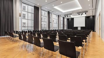 Uma sala de conferência com fileiras de cadeiras, janelas amplas e um palco com projetor.