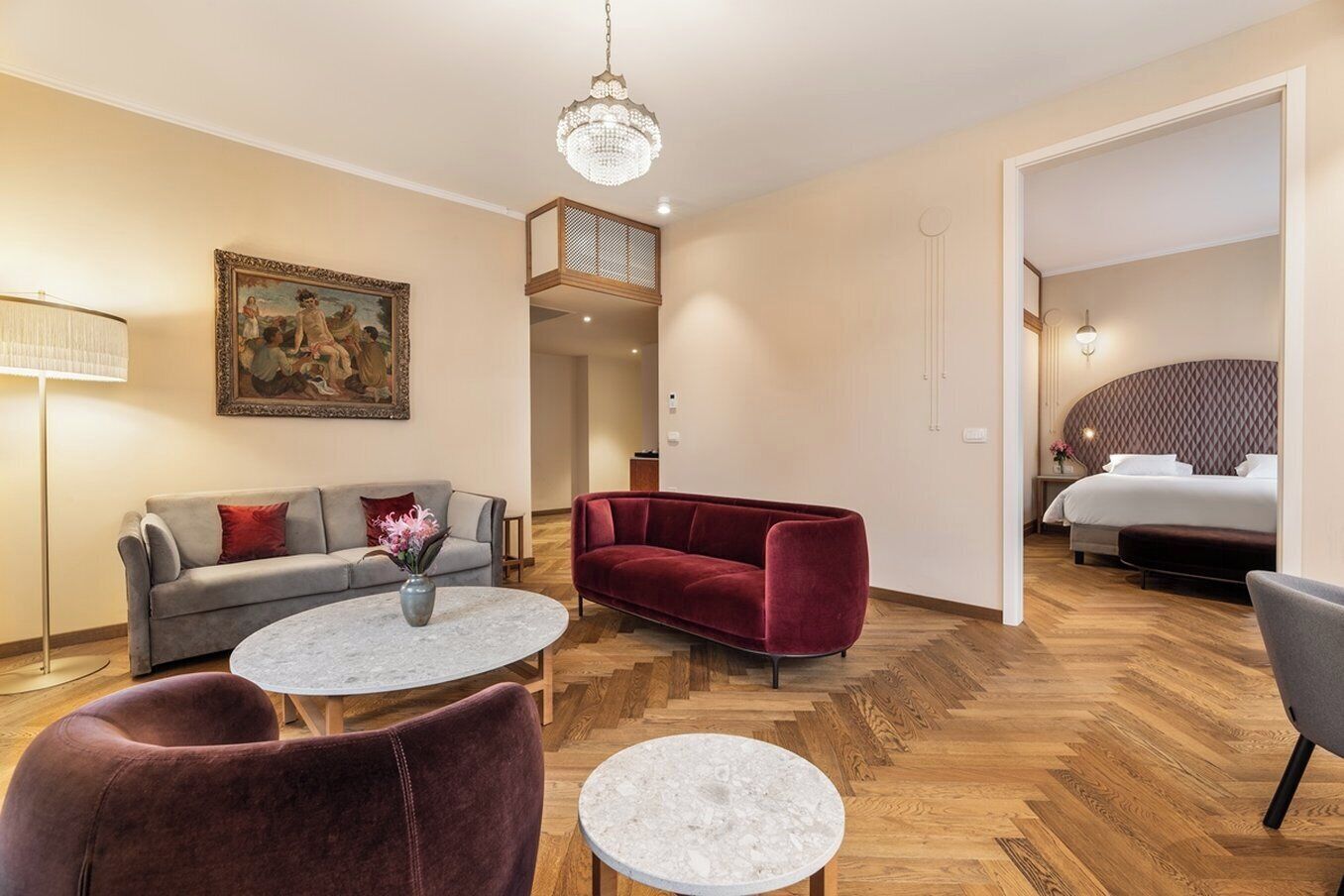 Uma sala de estar moderna com um sofá cinza, um sofá de veludo vermelho, uma mesa de centro de mármore e uma mesa redonda. Um candelabro pendurado no teto e um quadro na parede.