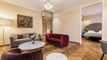 Uma sala de estar moderna com um sofá cinza, um sofá de veludo vermelho, uma mesa de centro de mármore e uma mesa redonda. Um candelabro pendurado no teto e um quadro na parede.