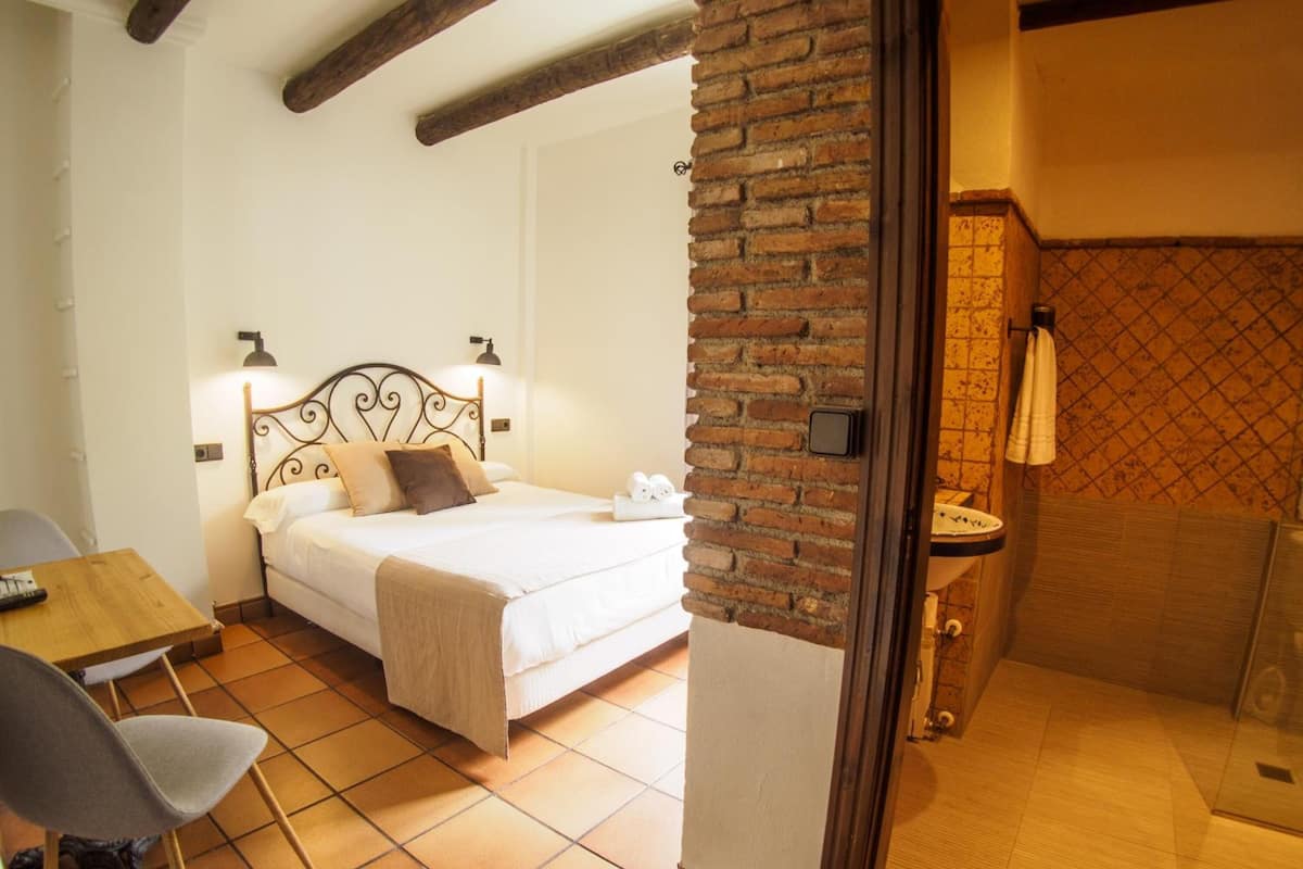Quarto com cama, mesa de cabeceira, cadeira e banheiro visível por uma porta aberta.