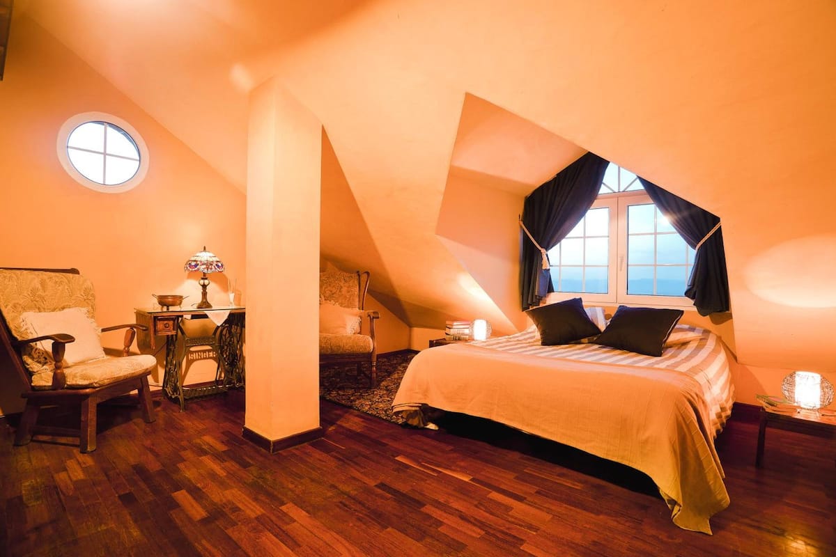 Quarto de sótão acolhedor com teto inclinado, uma cama grande, uma janela redonda e uma pequena mesa com abajur.
