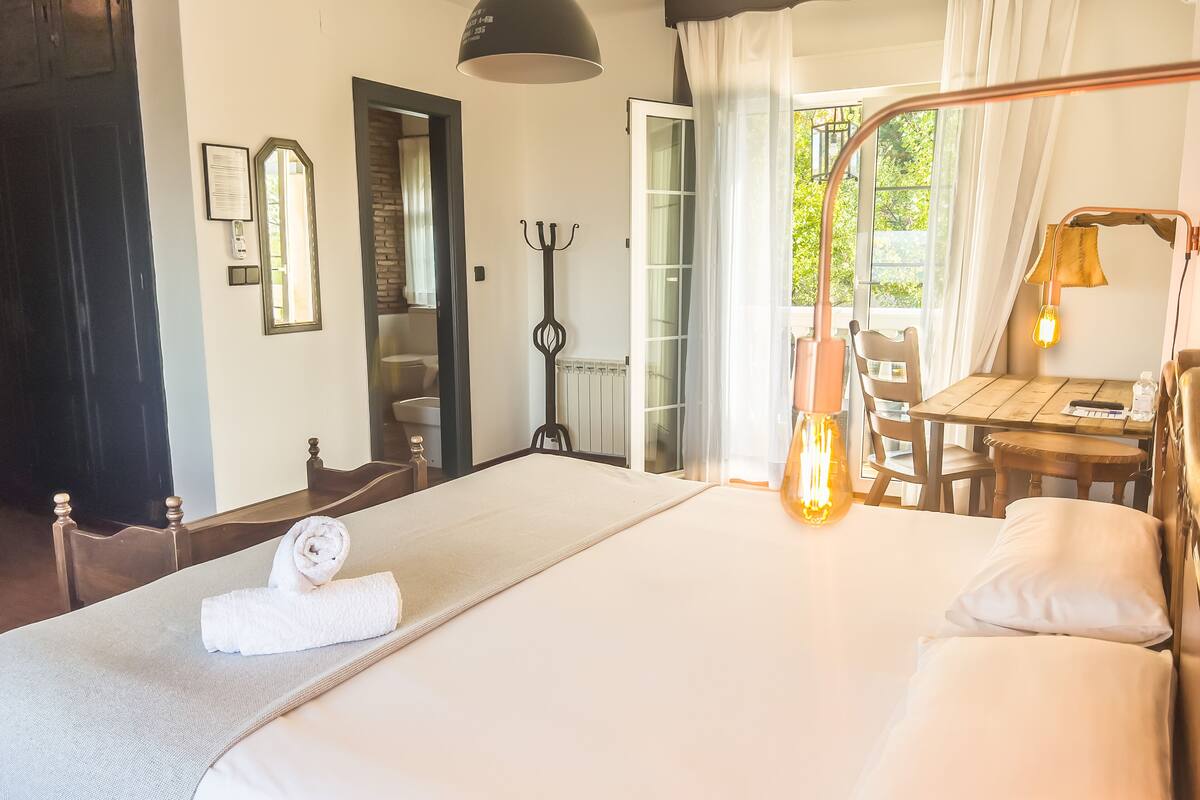 Quarto de hotel bem iluminado com uma cama grande, uma mesa de madeira e vista para o exterior pela janela.