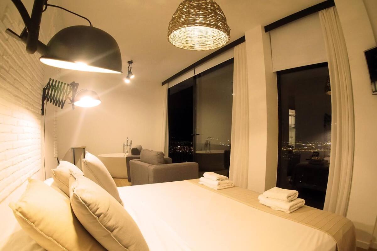 Quarto de hotel moderno com uma cama grande, área de estar, banheira e vista para a cidade.