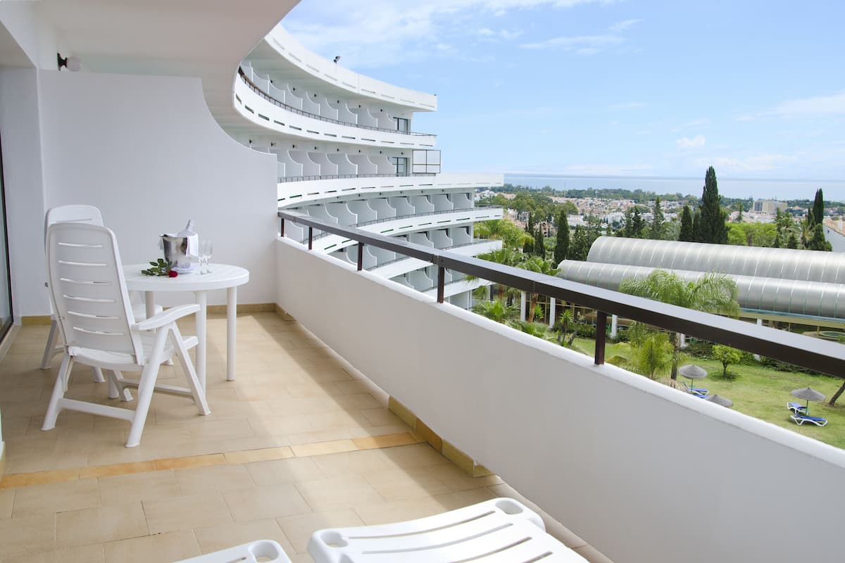 Varanda com mesa e cadeiras brancas, vista para a cidade e o mar, e um edifício moderno com varandas.
