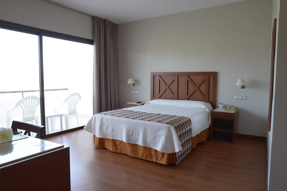 Quarto de hotel com uma cama grande, duas mesas de cabeceira, uma escrivaninha com cadeira e vista para o exterior através de uma janela ampla.