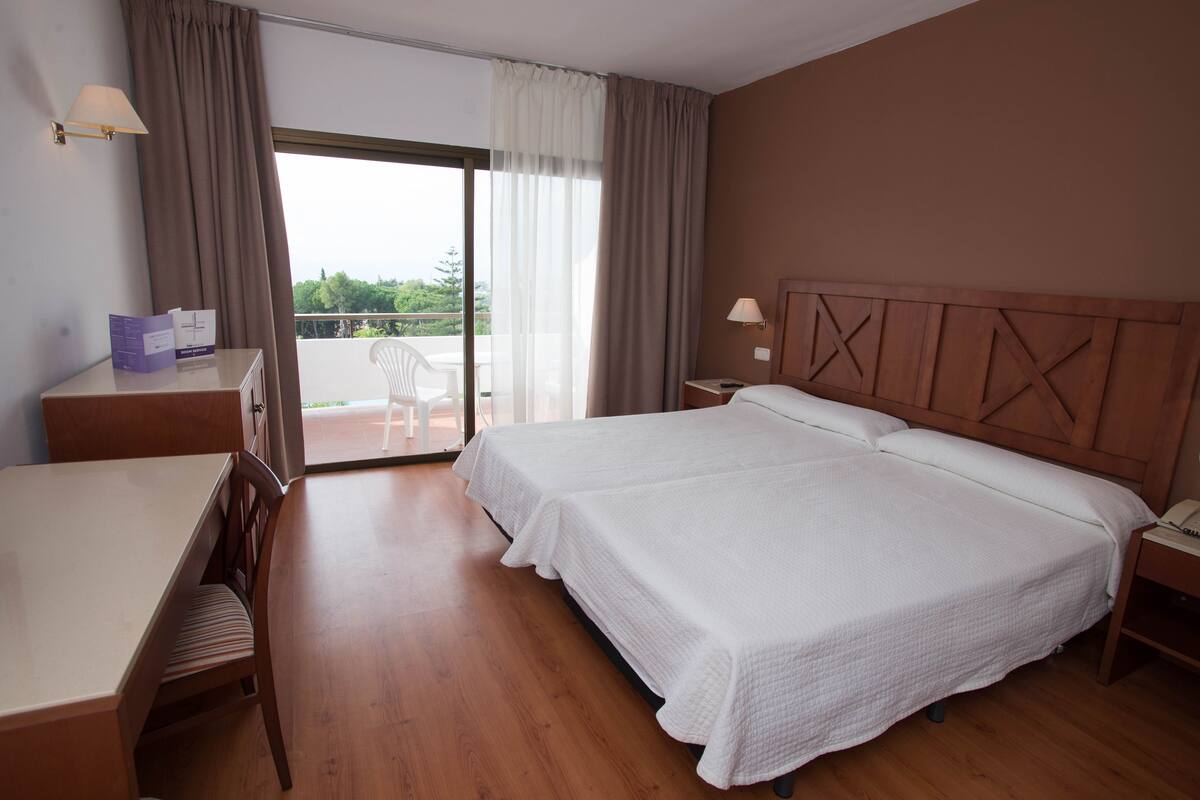 Quarto de hotel com uma cama grande, uma escrivaninha com cadeira, uma mesinha e vista para o exterior através de uma porta de correr de vidro.