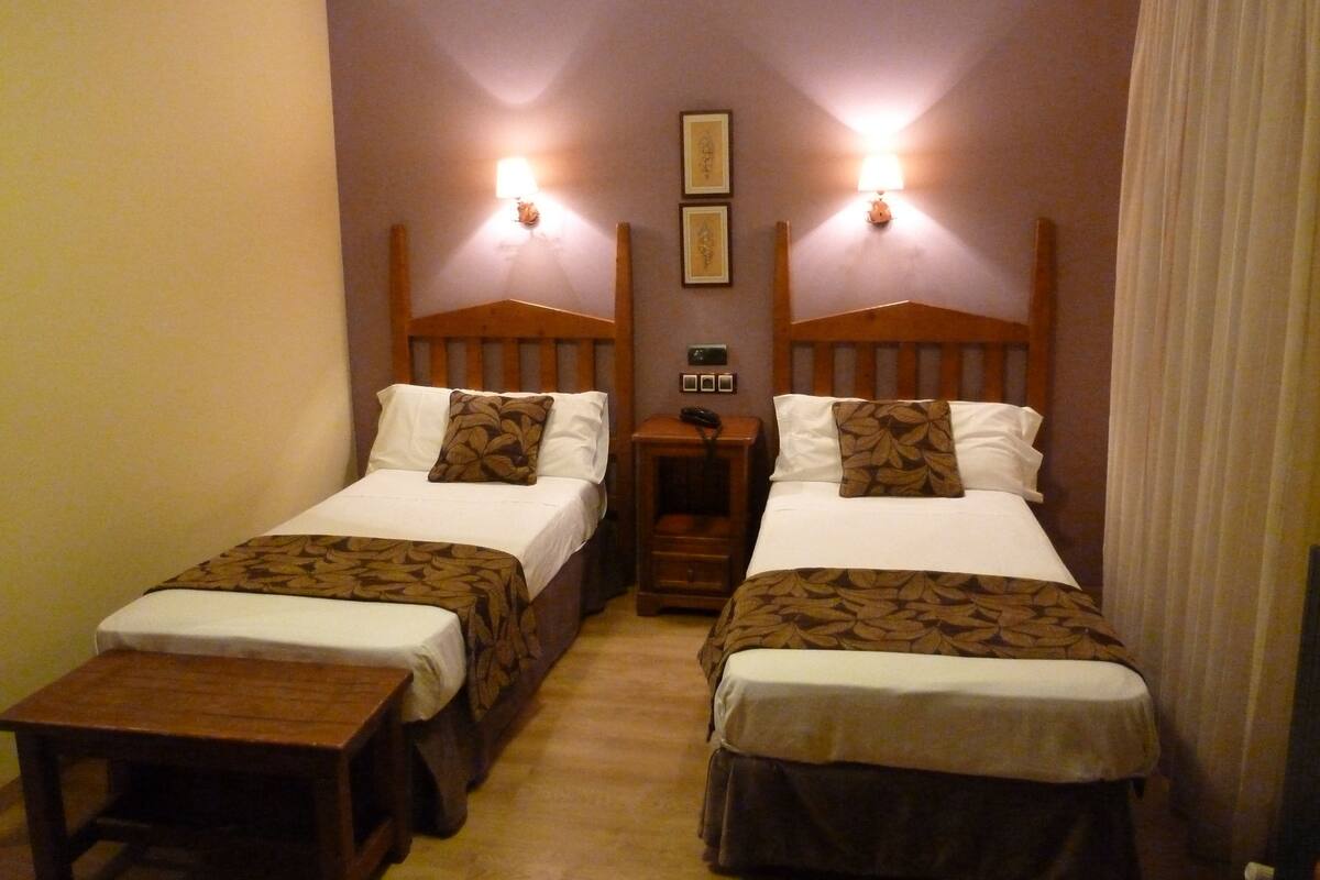 Quarto de hotel com duas camas de solteiro, cada uma com cabeceira de madeira, mesa de cabeceira e luminária.