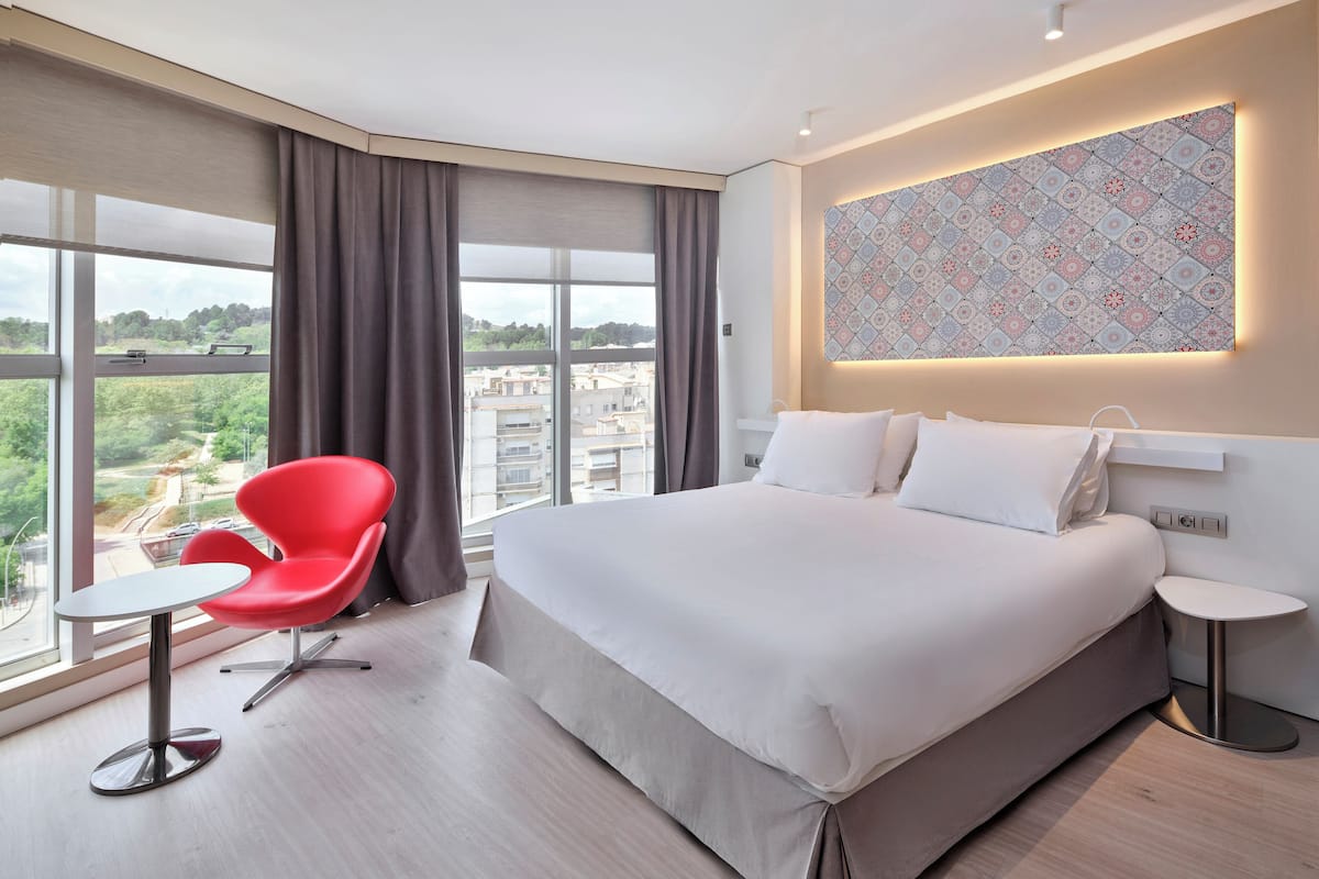 Quarto de hotel moderno com uma cama grande, uma cadeira vermelha, uma mesa pequena e vista da cidade através de amplas janelas.