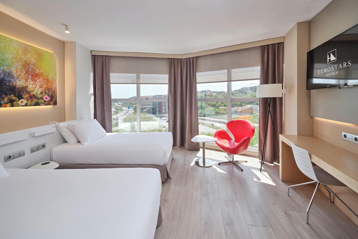 Quarto de hotel com duas camas, uma escrivaninha, televisão e uma grande janela com vista para a cidade.