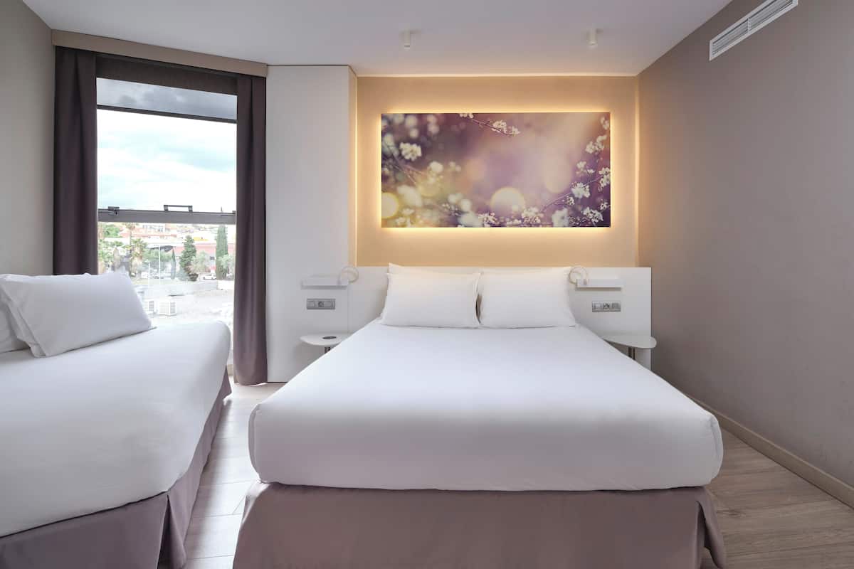 Quarto de hotel com uma cama grande, duas mesas de cabeceira, uma obra de arte na parede e uma janela com cortinas.