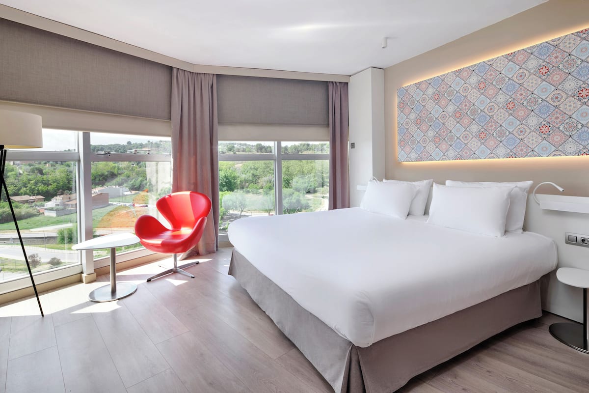 Quarto de hotel moderno com uma cama grande, uma cadeira vermelha, uma mesinha e vista para o exterior.