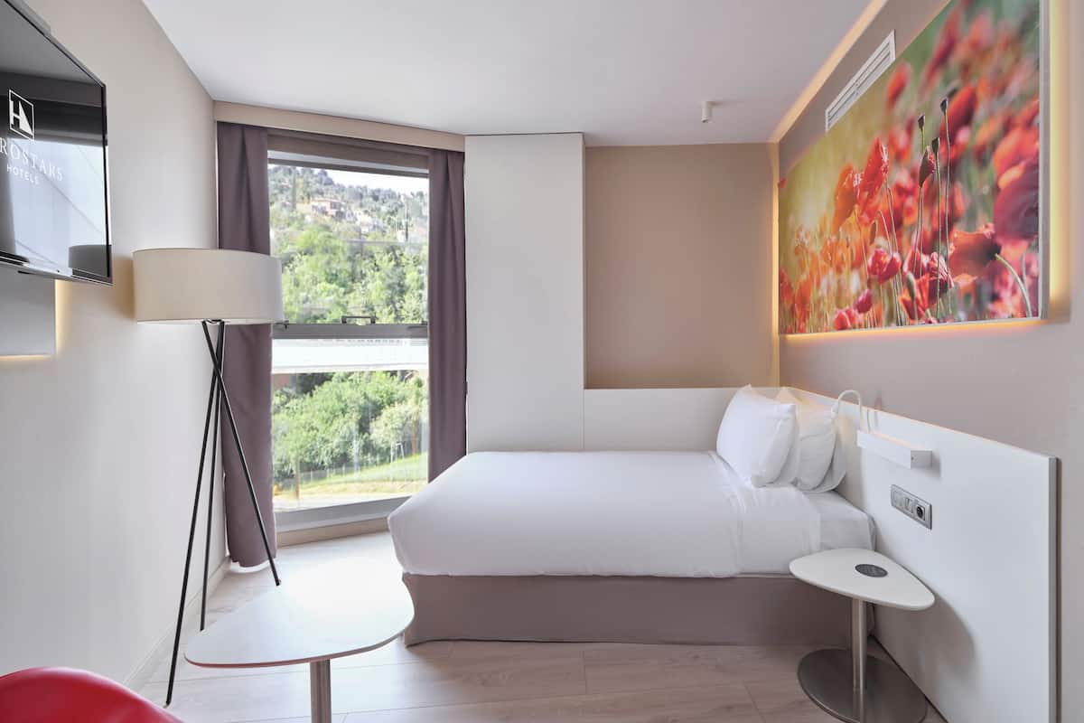 Quarto de hotel moderno com uma cama grande, uma mesinha, uma televisão e uma pintura de flores.