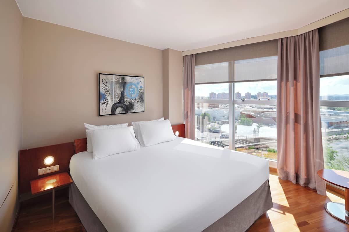 Quarto de hotel com uma cama grande, uma mesa de cabeceira, uma obra de arte na parede, uma janela com cortinas e vista para a cidade.