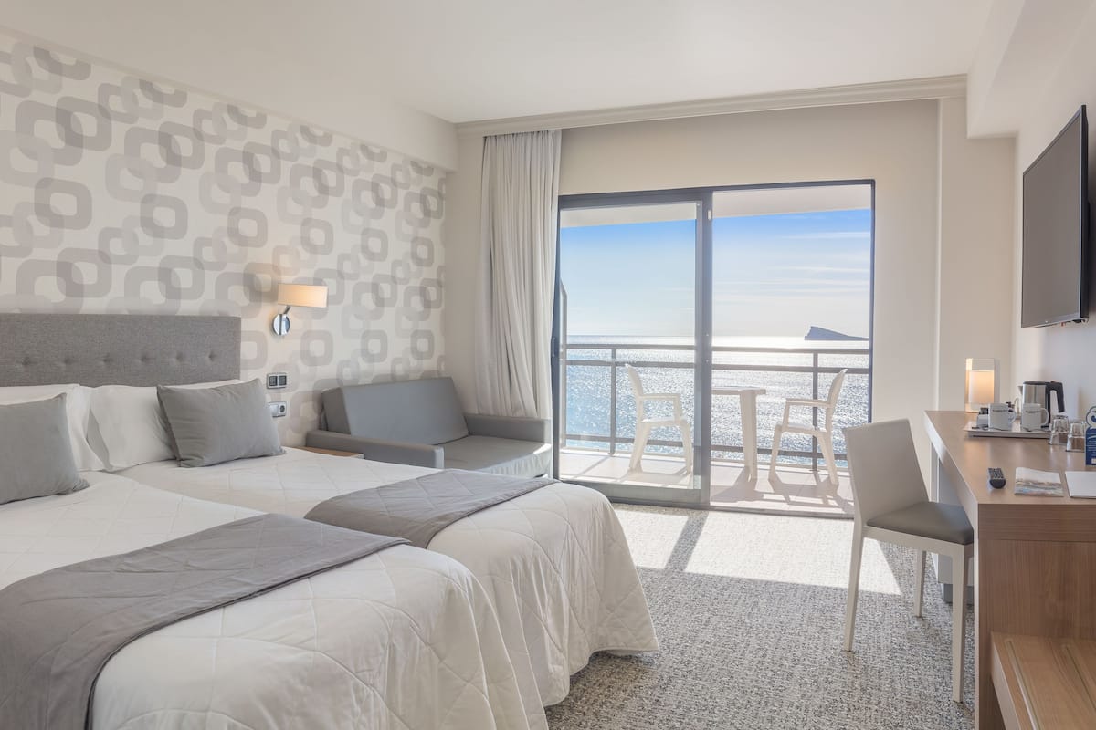 Quarto de hotel com duas camas, um sofá, uma mesa e uma varanda vista para o mar.