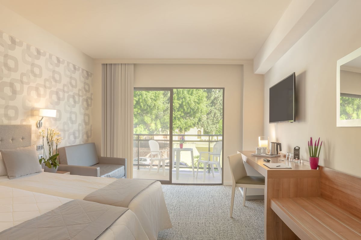 Quarto de hotel com duas camas, uma mesa, uma cadeira, televisão e varanda com vista para o verde.