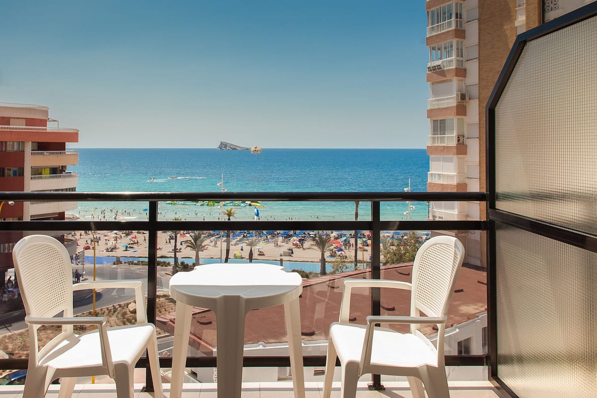Varanda com cadeiras e mesa brancas, com vista para uma praia com guarda-sóis e pessoas.