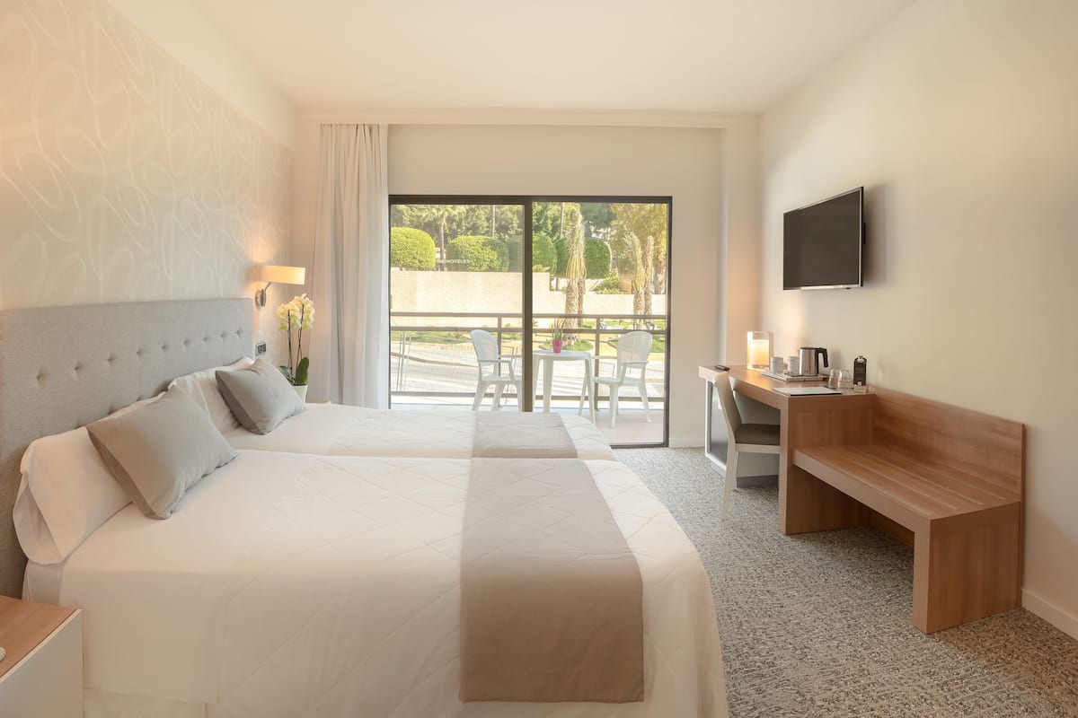 Quarto de hotel com duas camas, uma escrivaninha, televisão e varanda com vista para o verde.