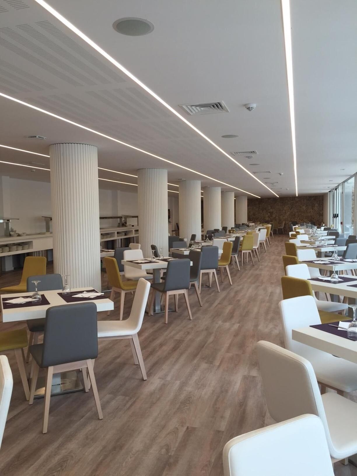 Uma área de jantar moderna, com mesas e cadeiras brancas, piso de madeira e uma fileira de colunas altas e brancas.