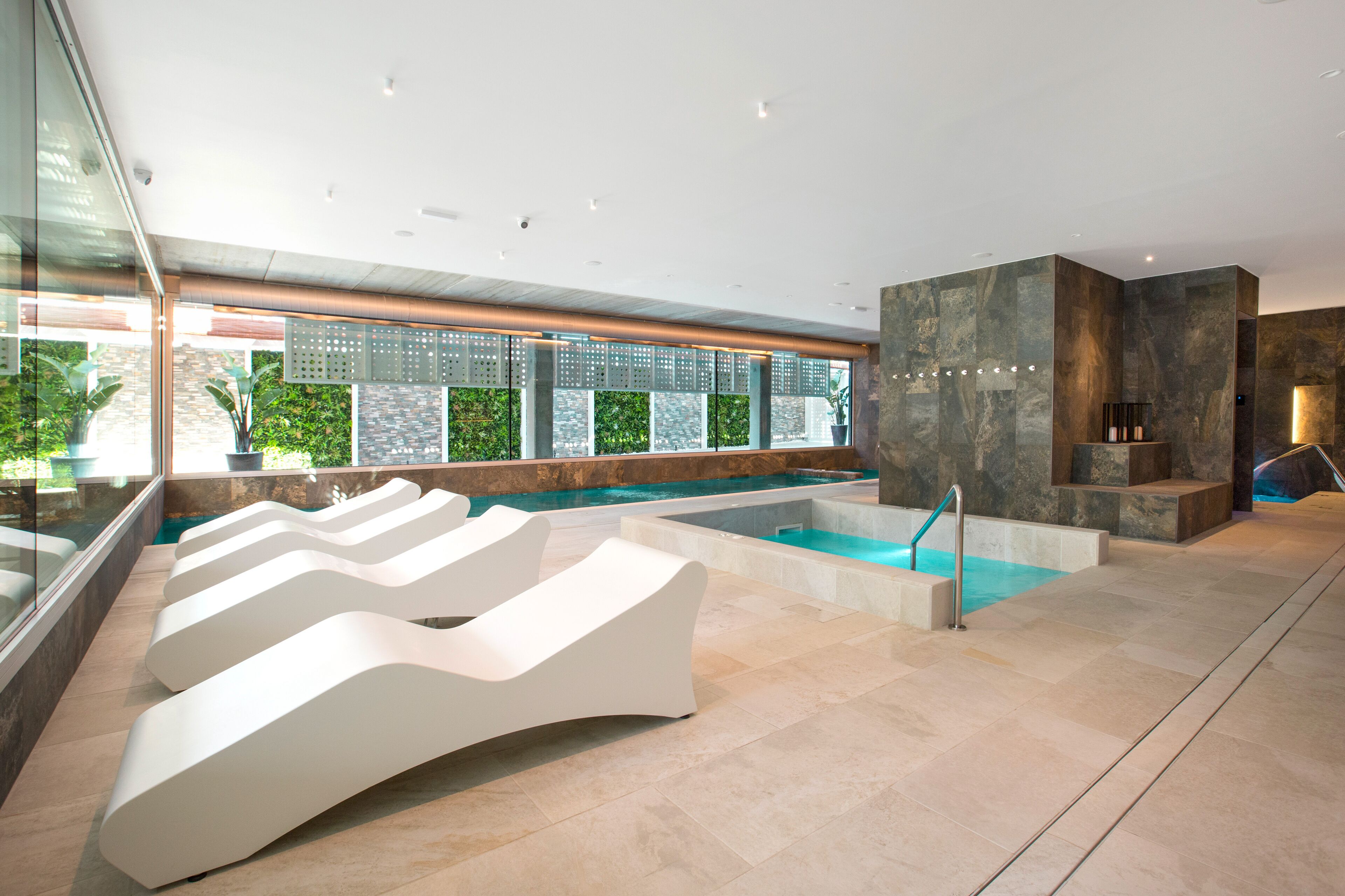 Uma área interna de piscina moderna, com parede de vidro, piscina com tonalidade azul e espreguiçadeiras brancas.
