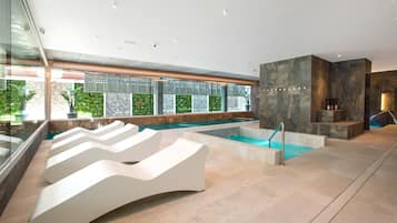 Uma área interna de piscina moderna, com parede de vidro, piscina com tonalidade azul e espreguiçadeiras brancas.