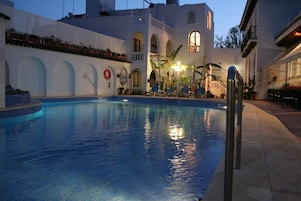 Piscina com superfície de água azul cristalina, cercada por um edifício branco com janelas em arco e varanda.