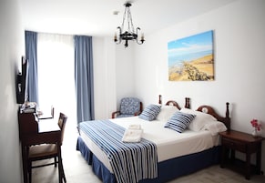 Quarto de hotel com cama, escrivaninha, cadeira, abajur, mesa de cabeceira, vaso com flores, pintura de uma praia e lustre.
