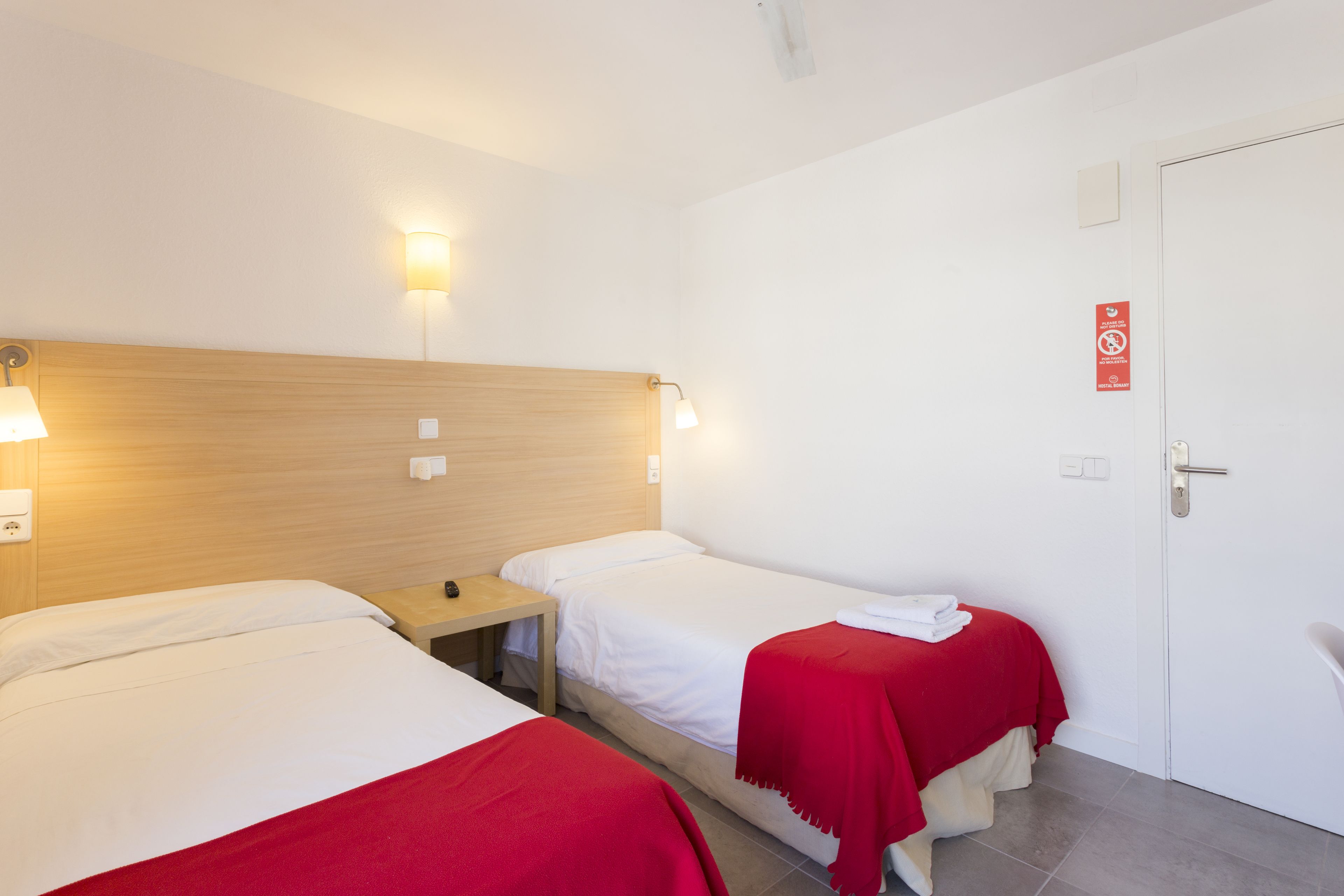 Quarto de hotel com duas camas, cabeceira de madeira, abajures, porta branca e um letreiro vermelho na parede.
