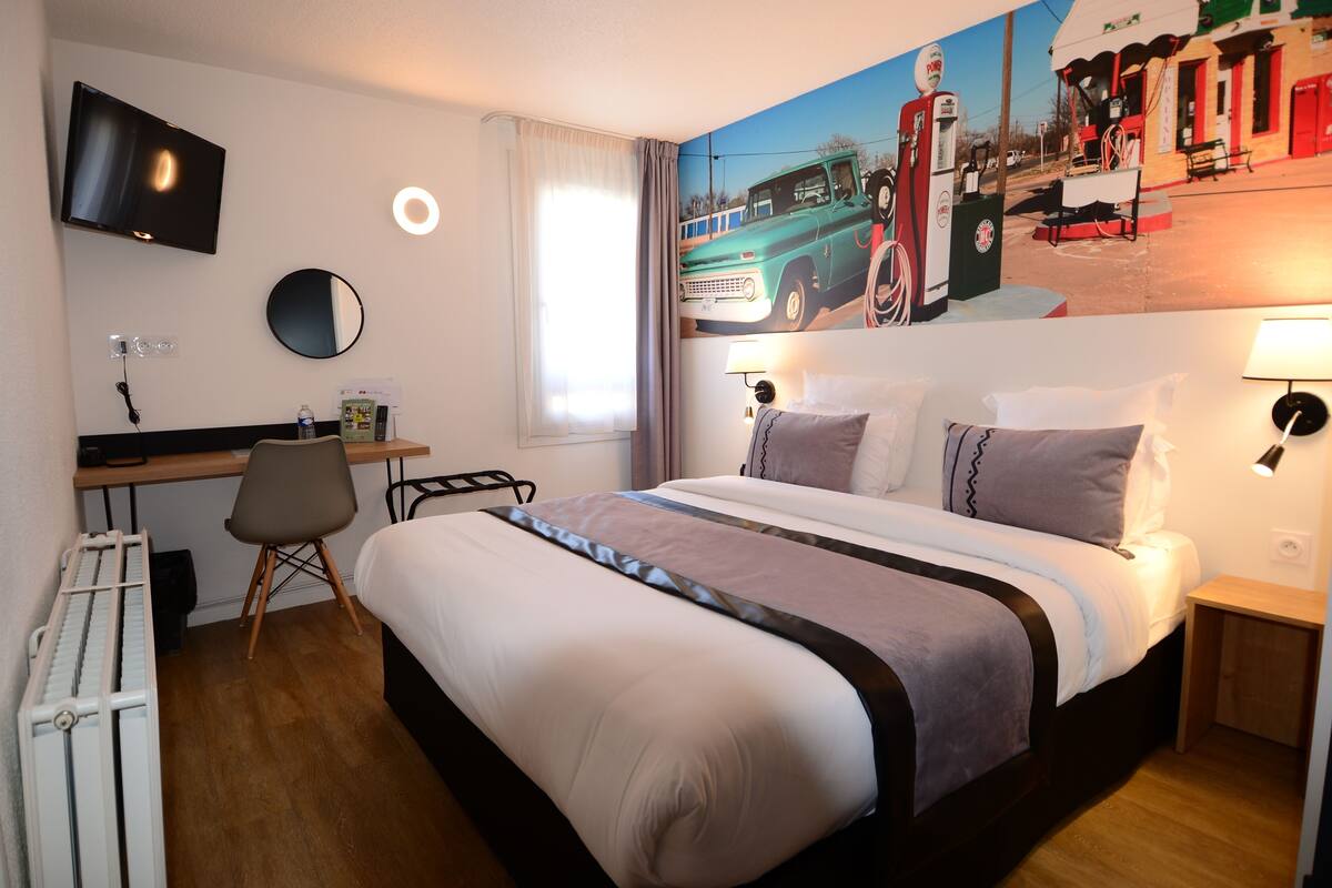 Quarto de hotel com cama, escrivaninha, cadeira e um quadro de um carro antigo e posto de gasolina.