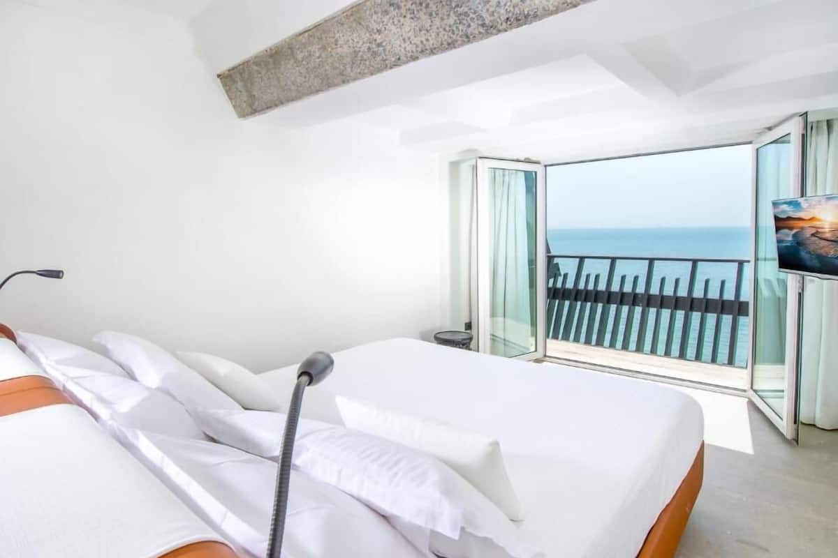 Quarto de hotel com cama, mesa de cabeceira, luminária e vista para o mar através de uma porta de vidro.