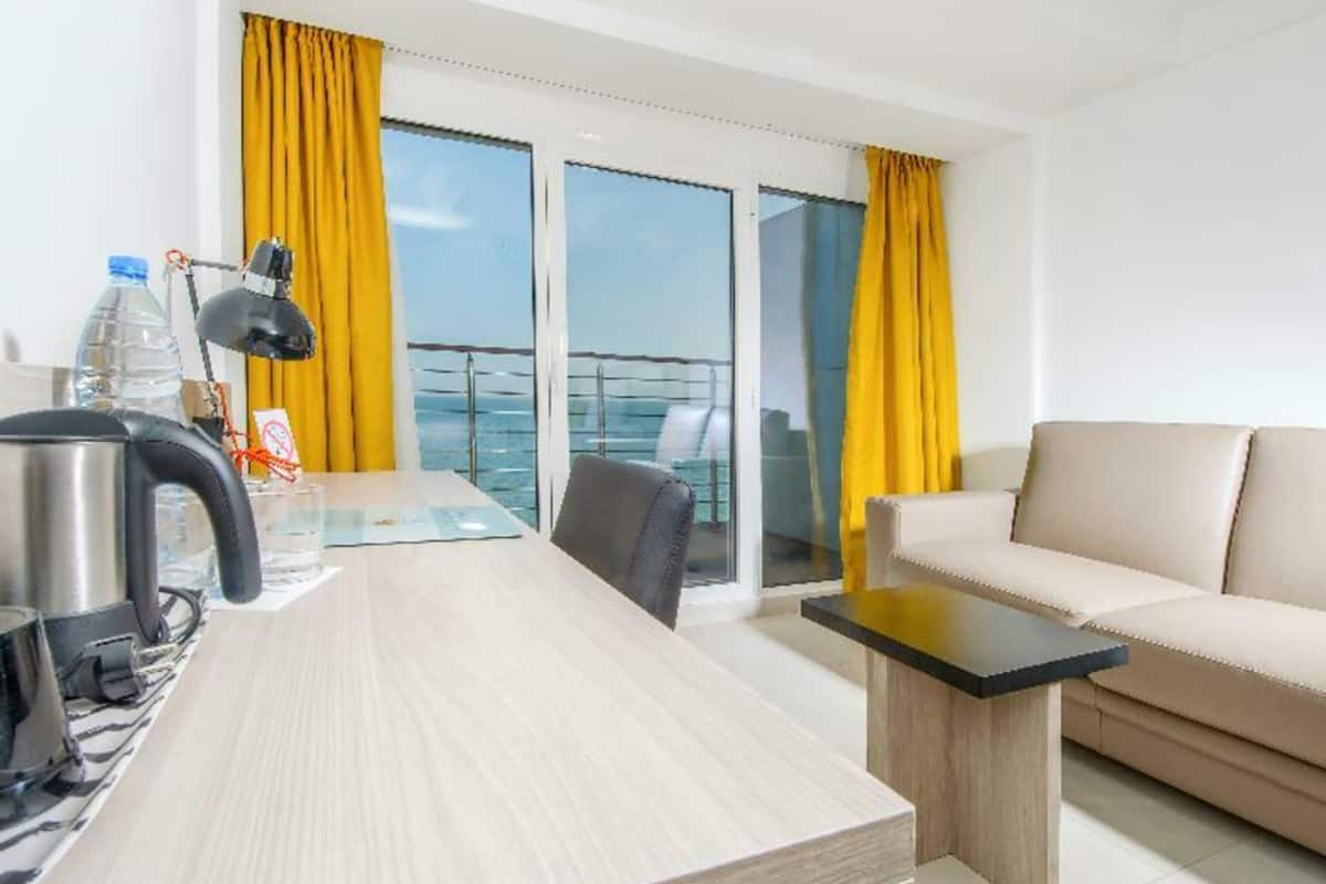 Quarto de hotel moderno com escrivaninha de madeira, sofá e varanda com cortinas amarelas.