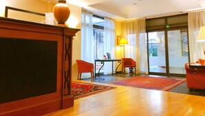 Um hall de hotel com um balcão de recepção de madeira, duas cadeiras vermelhas, uma mesa pequena com abajur e uma janela grande com cortinas.
