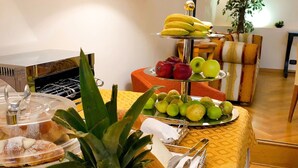 Mesa de buffet com diversas frutas e doces, uma luminária e uma planta em um quarto de hotel.