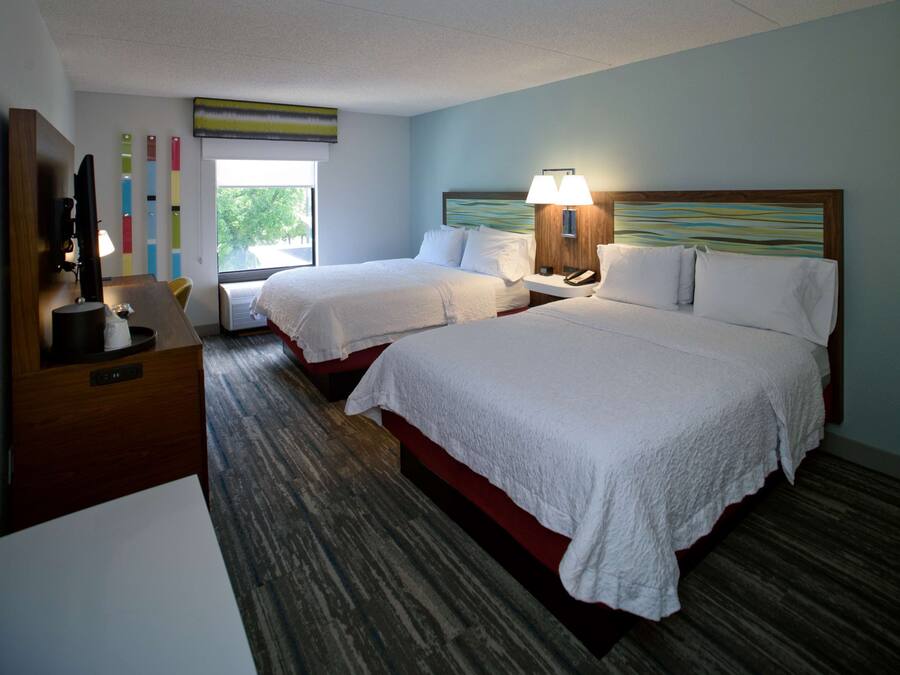 Quarto de hotel com duas camas, uma escrivaninha, uma cadeira, televisão e janela com vista para árvores.