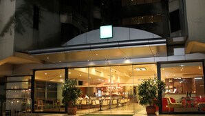 Entrada moderna de hotel com fachada de vidro, área de estar externa e plantas em vasos.