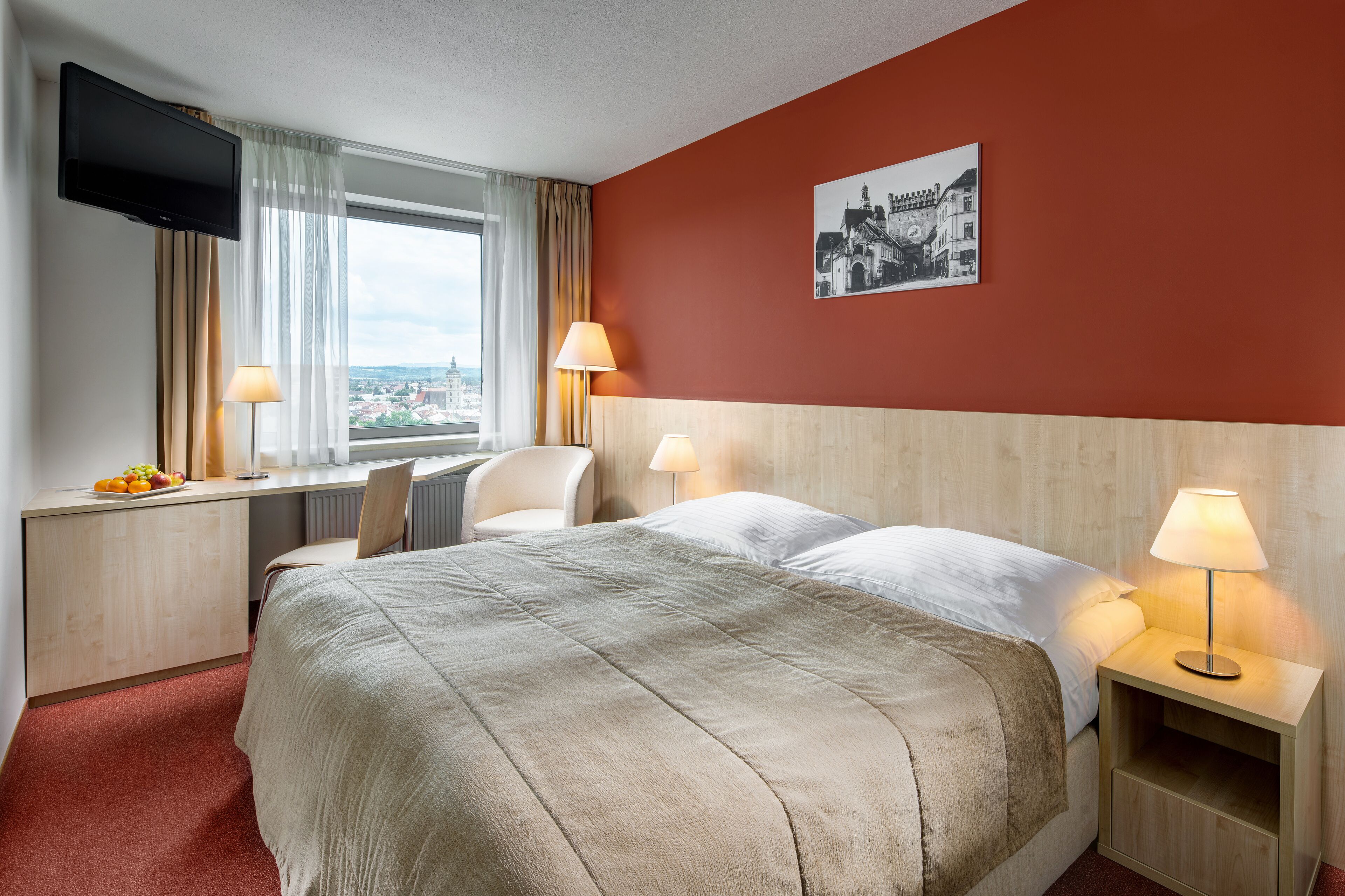 Quarto de hotel com cama, duas luminárias laterais, televisão, poltrona, escrivaninha com frutas e vista para a cidade.