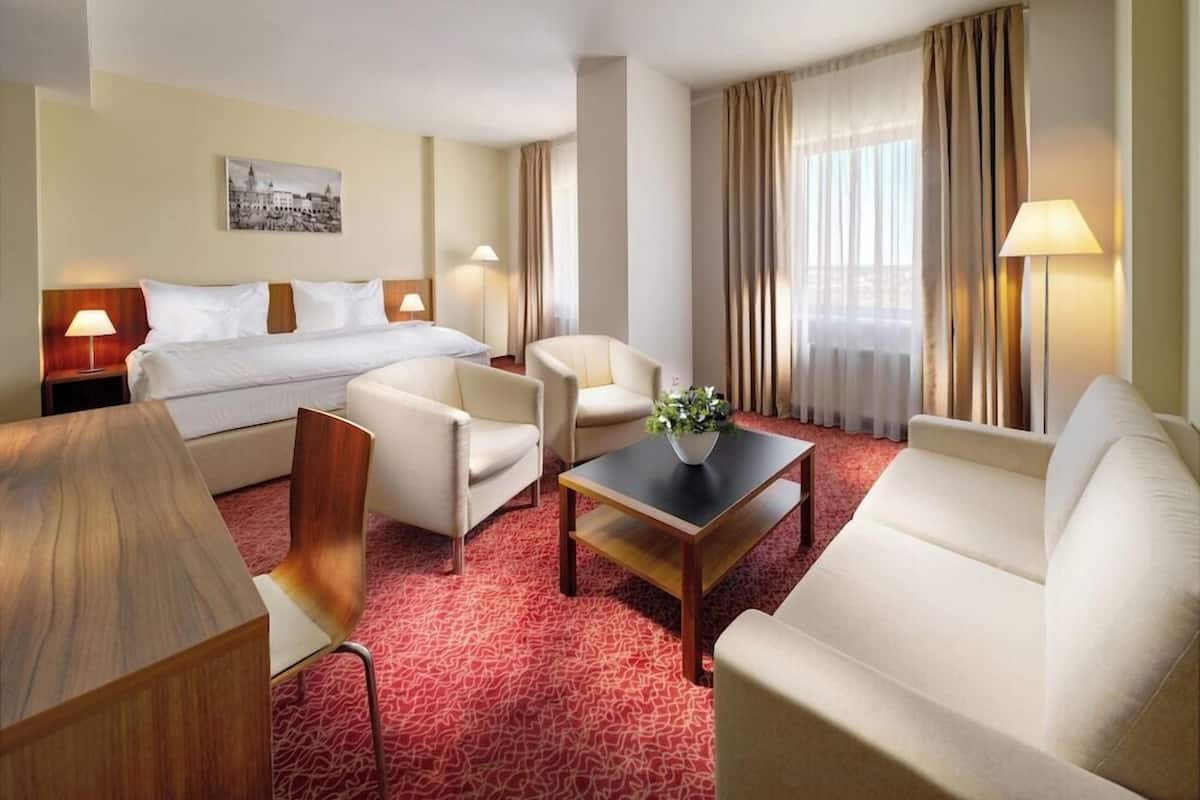 Quarto de hotel com cama, duas poltronas, um sofá, uma mesa de centro e um abajur.