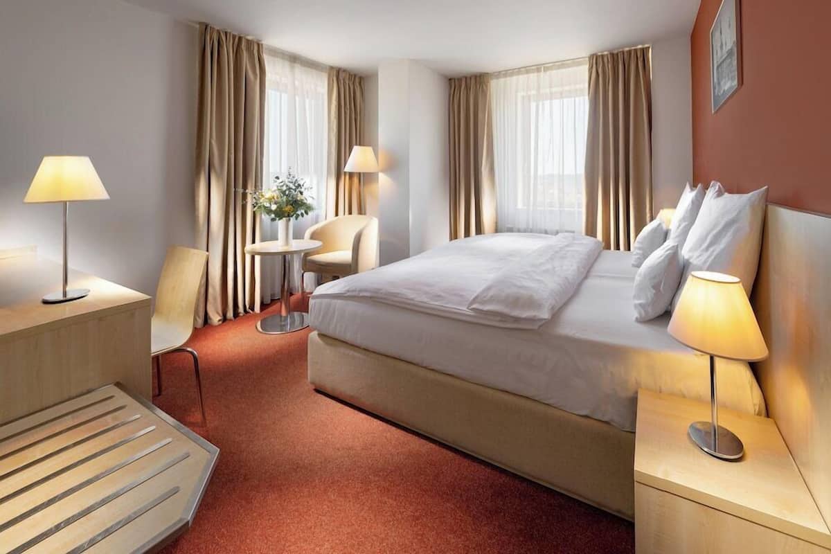 Quarto de hotel com uma cama, duas mesas de cabeceira com luminárias, uma cadeira, uma pequena mesa com um vaso e uma janela com cortinas.