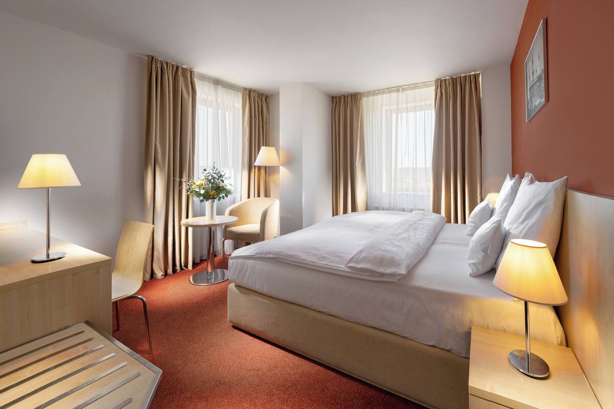 Quarto de hotel com uma cama grande, duas mesas de cabeceira com luminárias, uma cadeira, uma mesinha com um vaso e um quadro na parede.