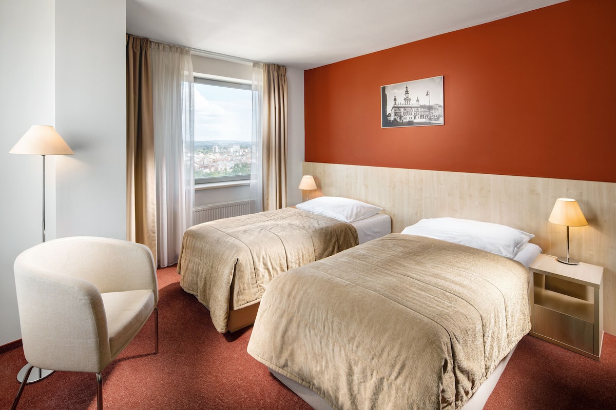 Quarto de hotel com duas camas, uma cadeira, uma mesa de cabeceira, uma luminária e uma janela com cortinas.