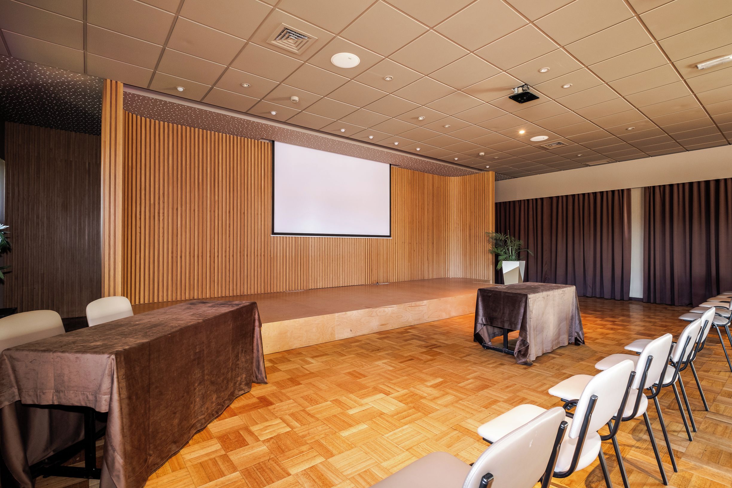 Sala de conferências com palco, tela de projetor, painel de madeira e uma fileira de cadeiras.