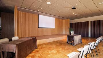 Sala de conferências com palco, tela de projetor, painel de madeira e uma fileira de cadeiras.