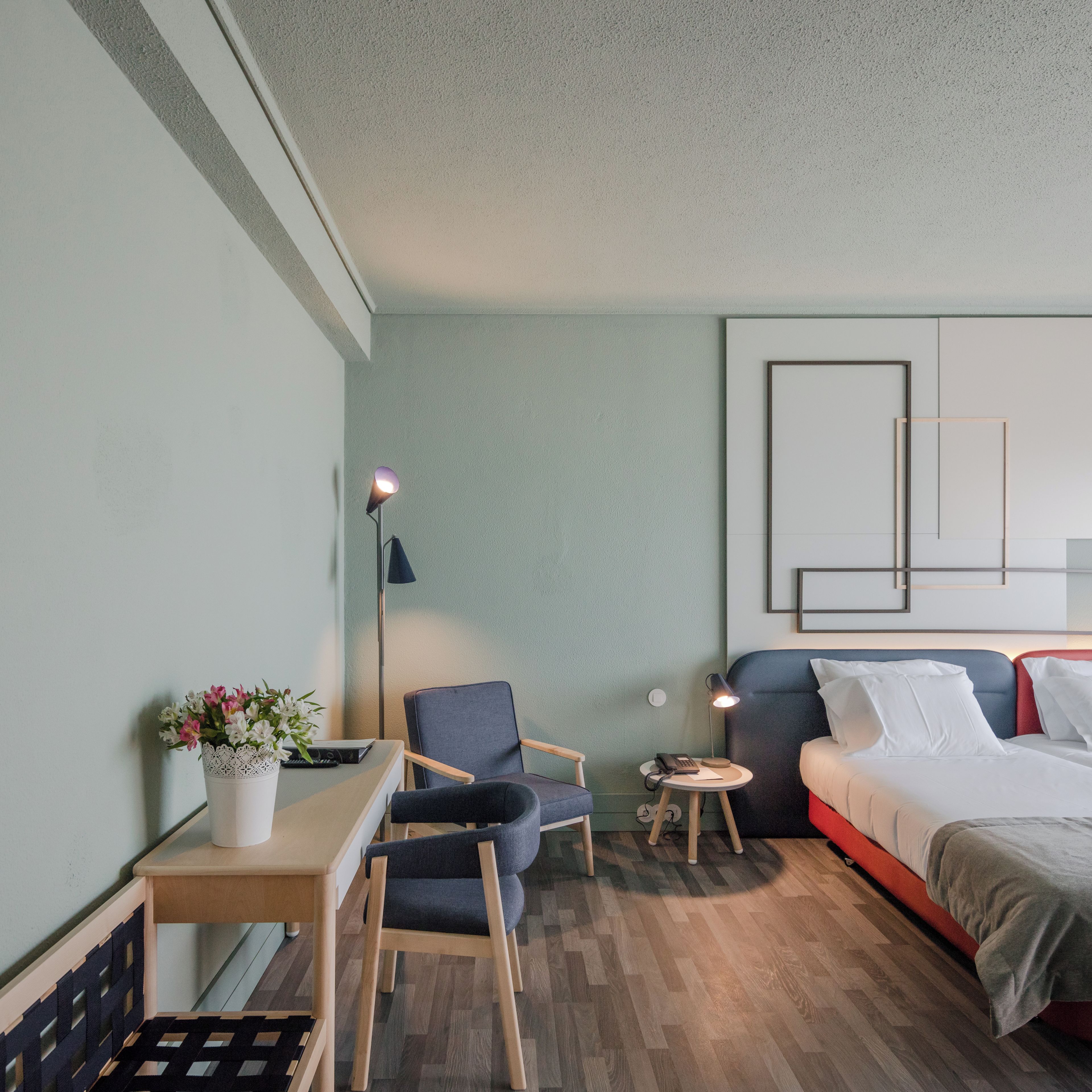 Quarto de hotel com cama, escrivaninha, cadeira, mesinha e abajur.