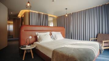 Quarto de hotel com uma cama grande, uma mesa de cabeceira com luminária, uma cadeira e janela com cortina.