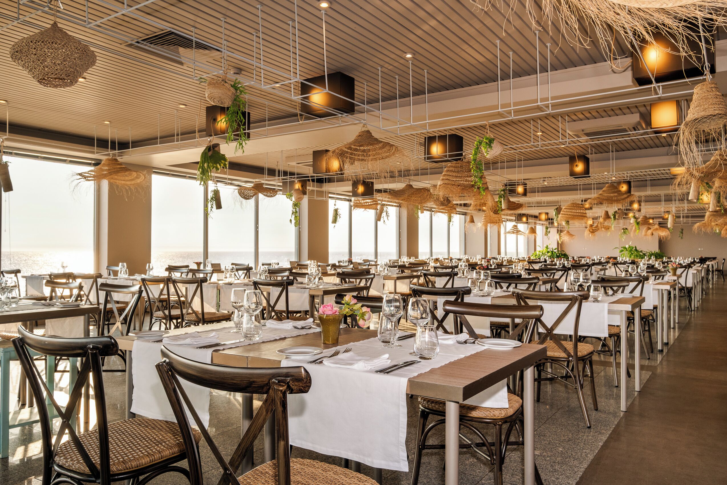 Um restaurante amplo com várias mesas e cadeiras, janelas grandes e plantas penduradas.