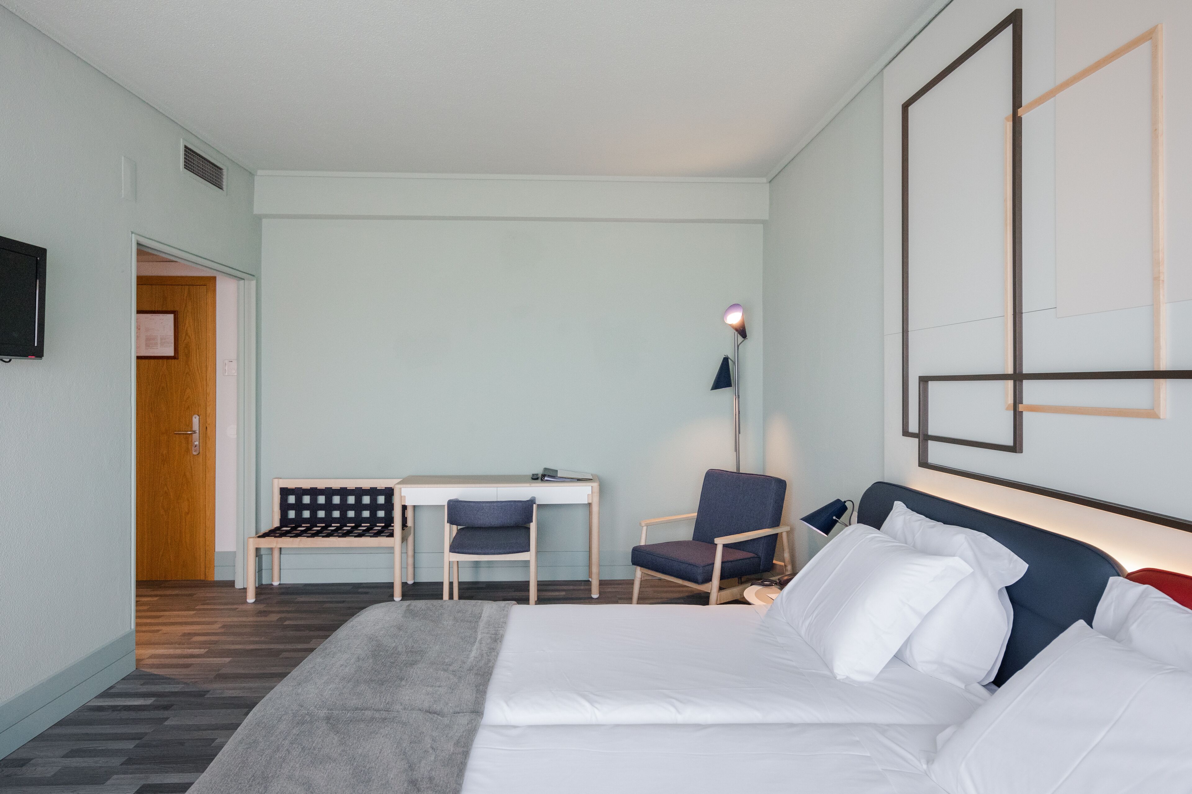 Quarto de hotel com cama, escrivaninha, duas cadeiras e televisão na parede.