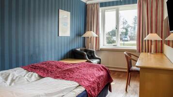 Quarto com cama, escrivaninha, duas cadeiras, janela com cortinas e radiador.