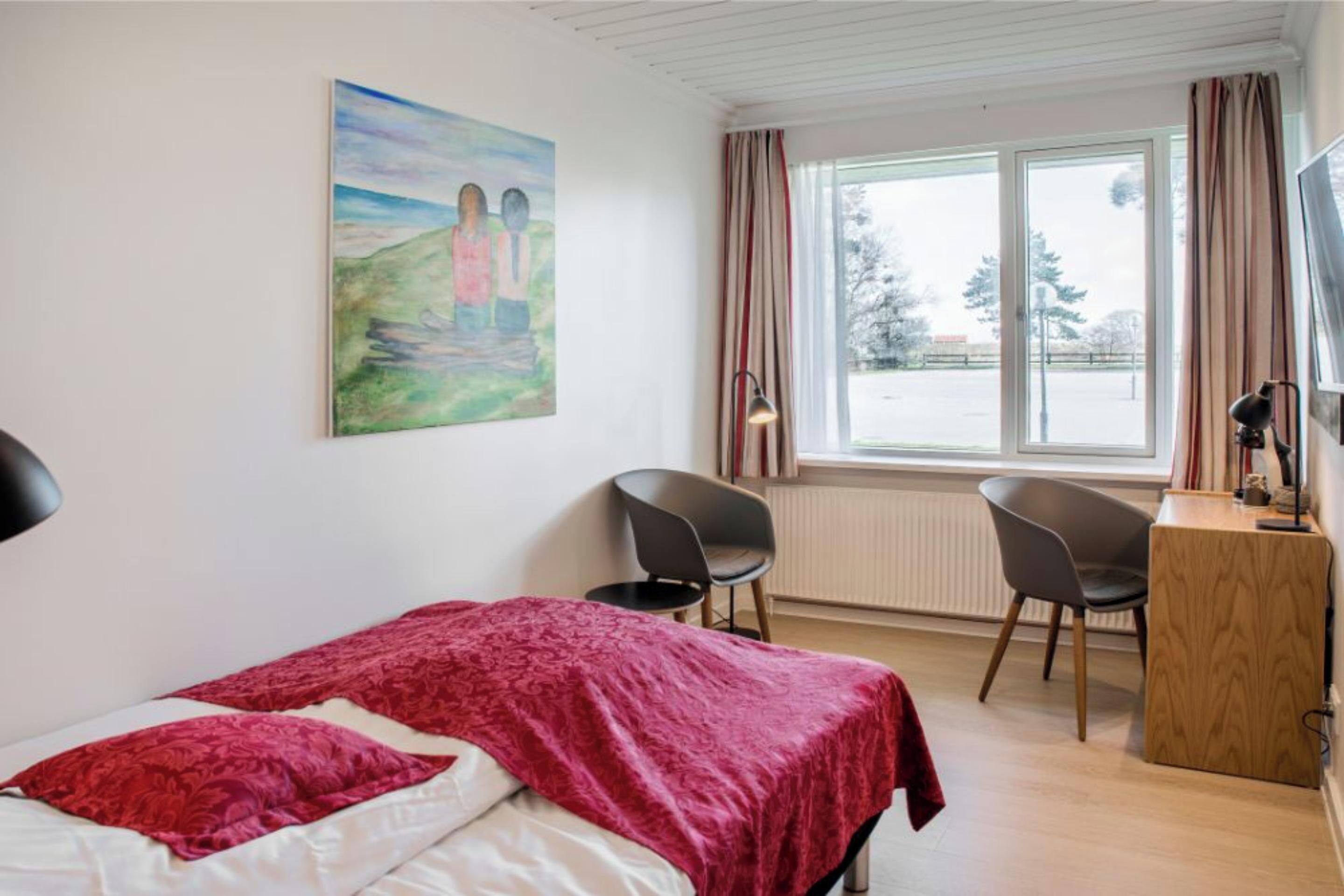 Quarto com cama, escrivaninha, duas cadeiras, radiador e janela com cortinas.
