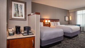 Quarto de hotel com duas camas, uma cozinha compacta, um quadro na parede e uma janela com cortinas.