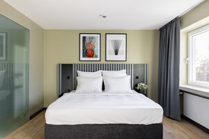 Quarto de hotel moderno com uma cama grande, cabeceira com linhas verticais, duas obras de arte emolduradas na parede e uma janela com cortinas.