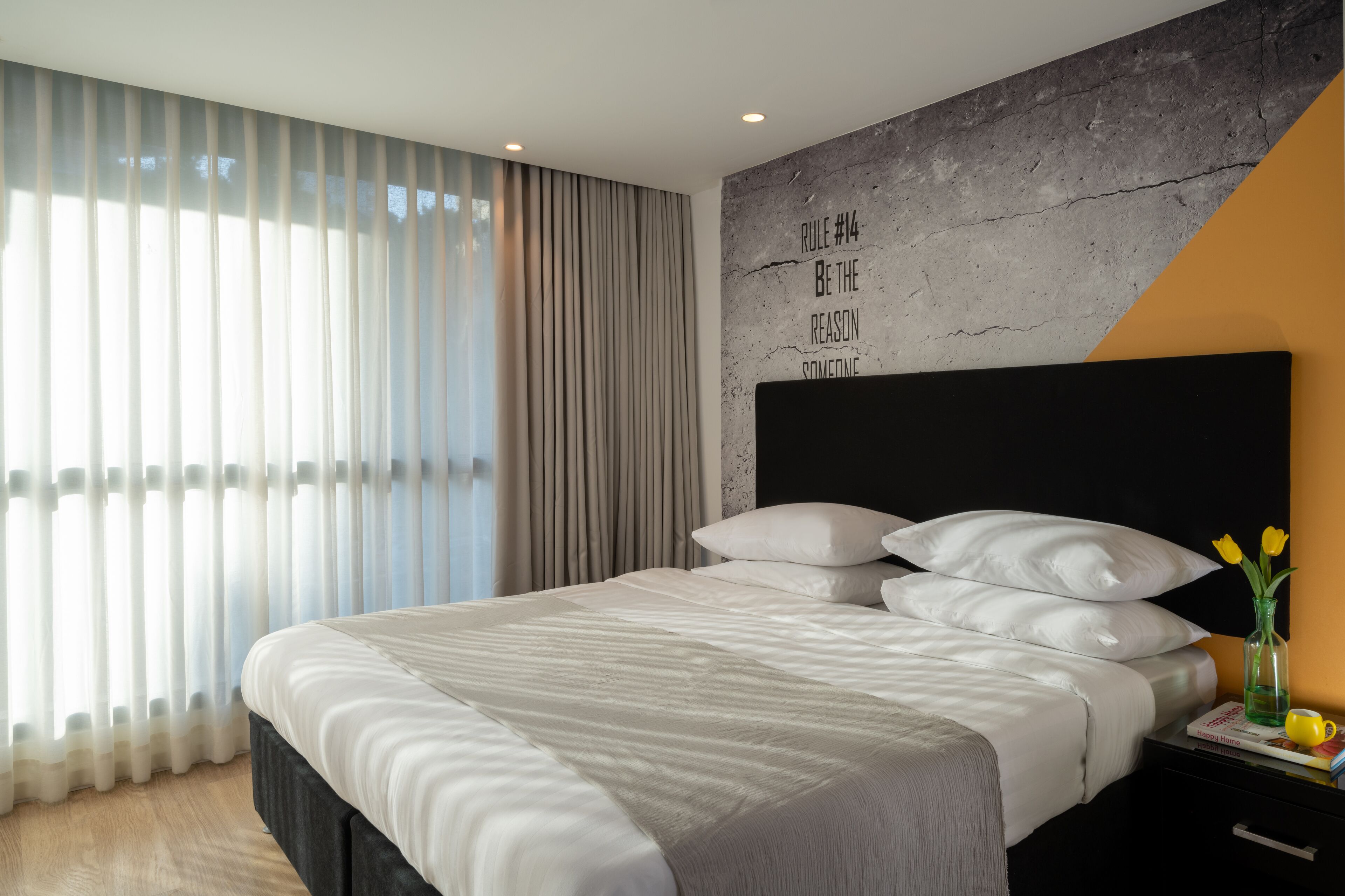 Quarto de hotel moderno com uma cama grande, roupa de cama branca, cabeceira preta, mesa de cabeceira com um vaso de flores e uma frase motivacional na parede.
